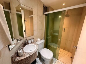 Casa de banho com bancada de granito, lavatório, sanita e cabine de duche com portas de vidro.