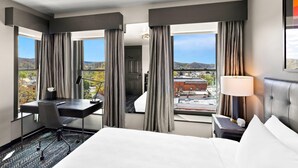 Quarto de hotel com cama, escrivaninha e cadeira, janelas amplas com cortinas, mesa de cabeceira com abajur e vista para a cidade.