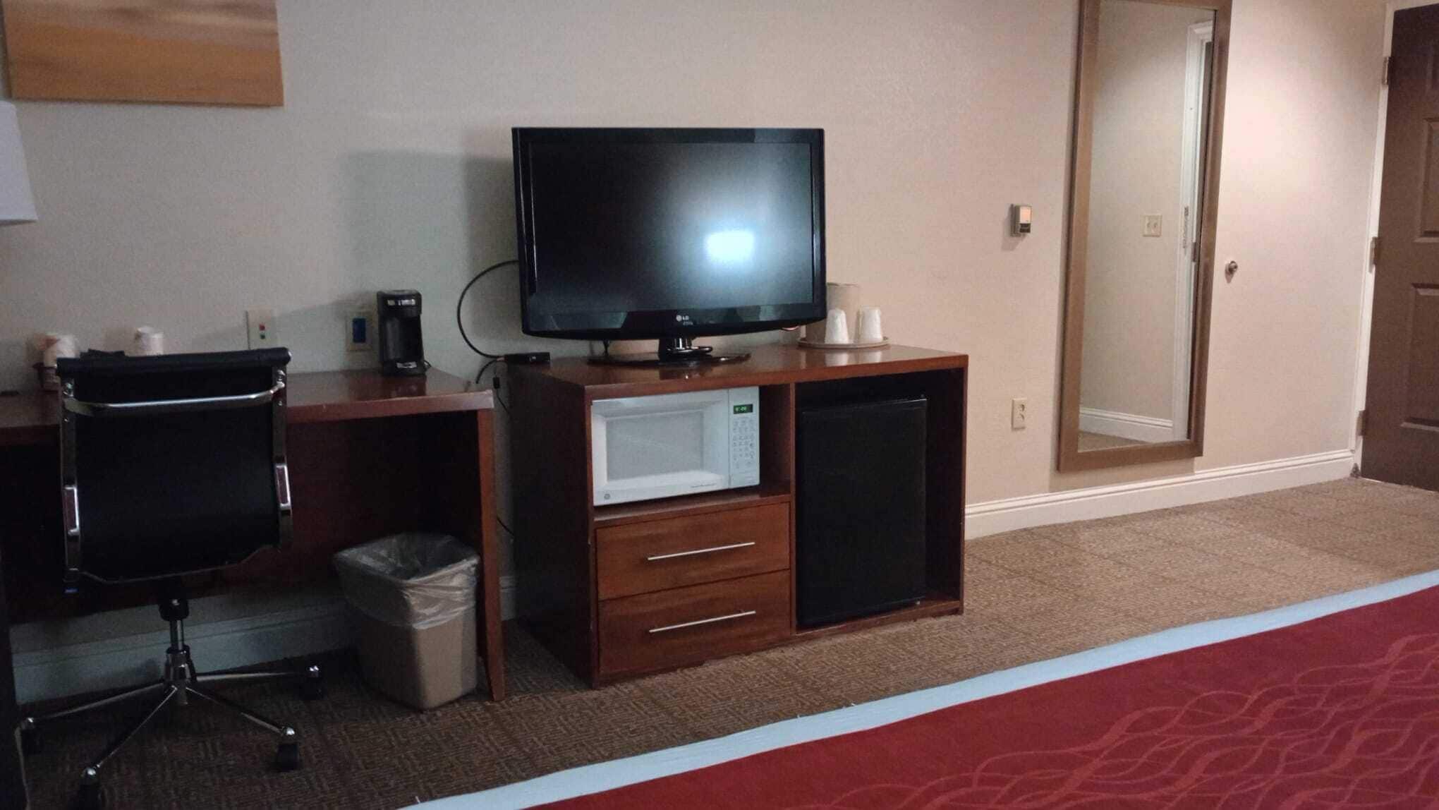 Quarto de hotel com escrivaninha, televisão, micro-ondas, lixeira e uma cadeira.