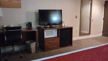 Quarto de hotel com escrivaninha, televisão, micro-ondas, lixeira e uma cadeira.