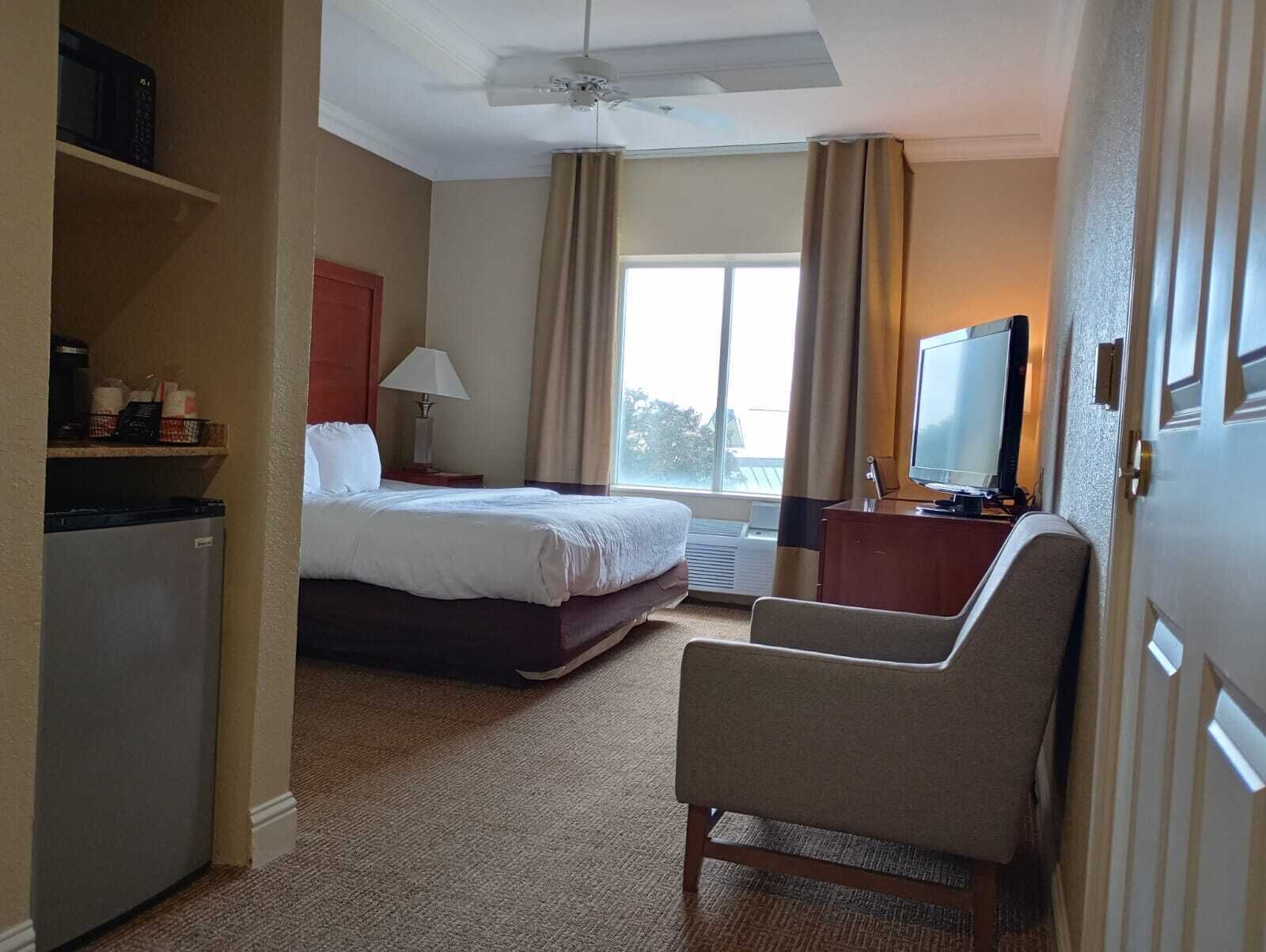 Quarto de hotel com uma cama, uma escrivaninha com computador, uma cadeira, televisão, ventoinha no teto e janela com cortinas.