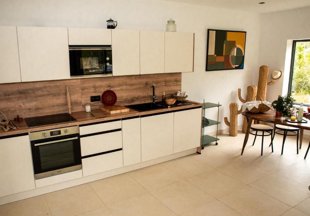 Cozinha moderna com armários brancos, forno embutido, micro-ondas e painel de madeira. Há uma área de refeições com mesa e cadeiras, e uma janela grande que oferece iluminação natural.