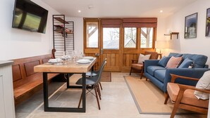 Uma sala de jantar com uma mesa de madeira posta para quatro pessoas, um sofá e uma televisão na parede.