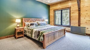 Quarto com cama de madeira, duas mesas de cabeceira com abajures, radiador e janela com vista para árvores.