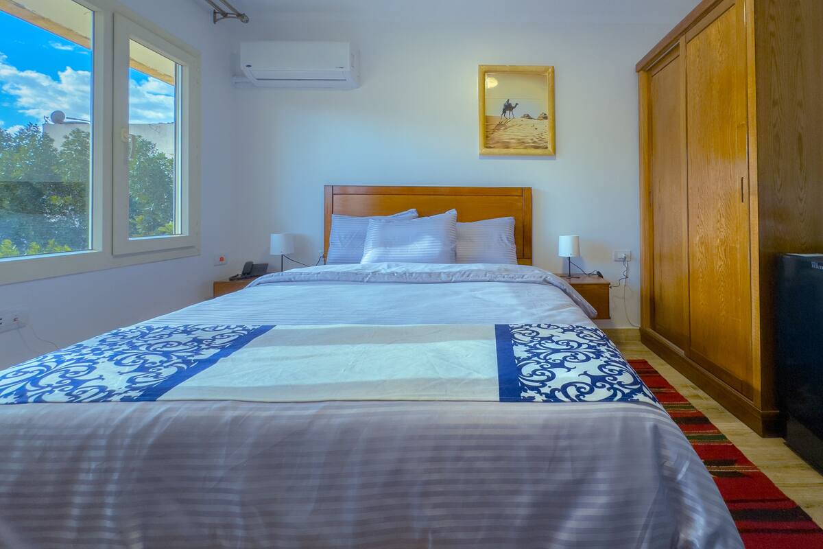 Uma cama bem arrumada com colcha estampada, cabeceira de madeira, abajures e vista para o exterior através de uma janela.