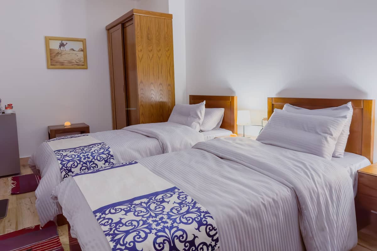 Quarto de hotel com duas camas, um guarda-roupa, uma mesinha de cabeceira com abajur e um quadro na parede.