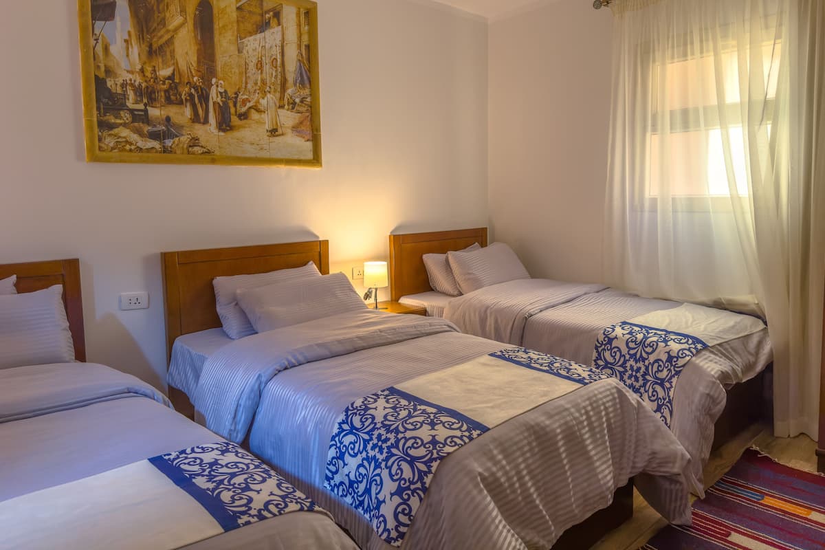 Quarto de hotel com três camas, um quadro na parede e uma janela com cortinas leves.