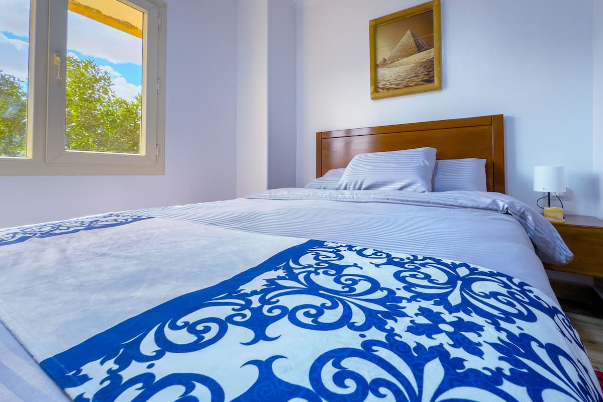 Cama bem arrumada com colcha estampada, cabeceira de madeira, mesa de cabeceira com abajur e quadro na parede.
