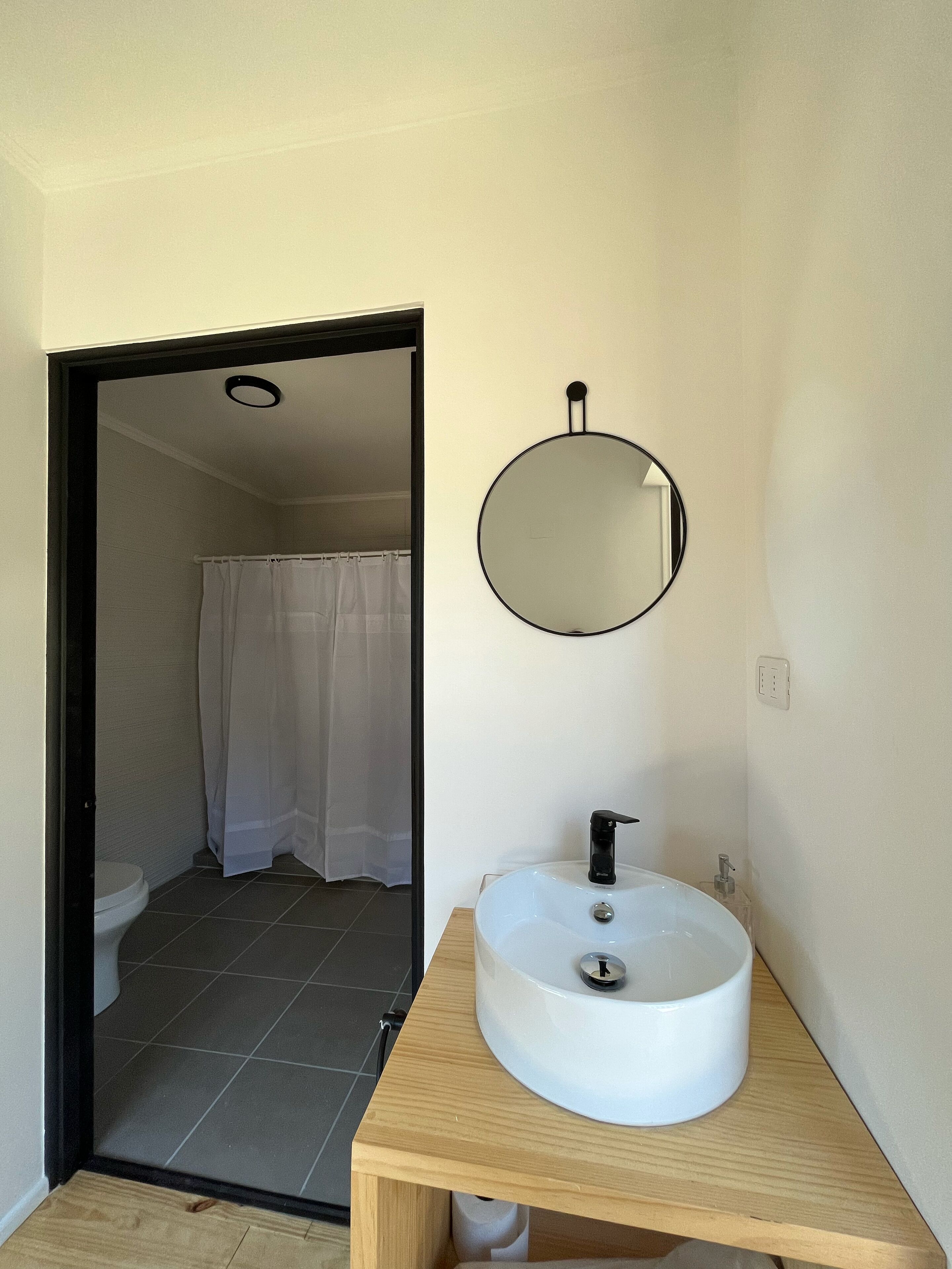 Casa de banho com lavatório branco, espelho redondo e zona de duche visível através de uma porta aberta.