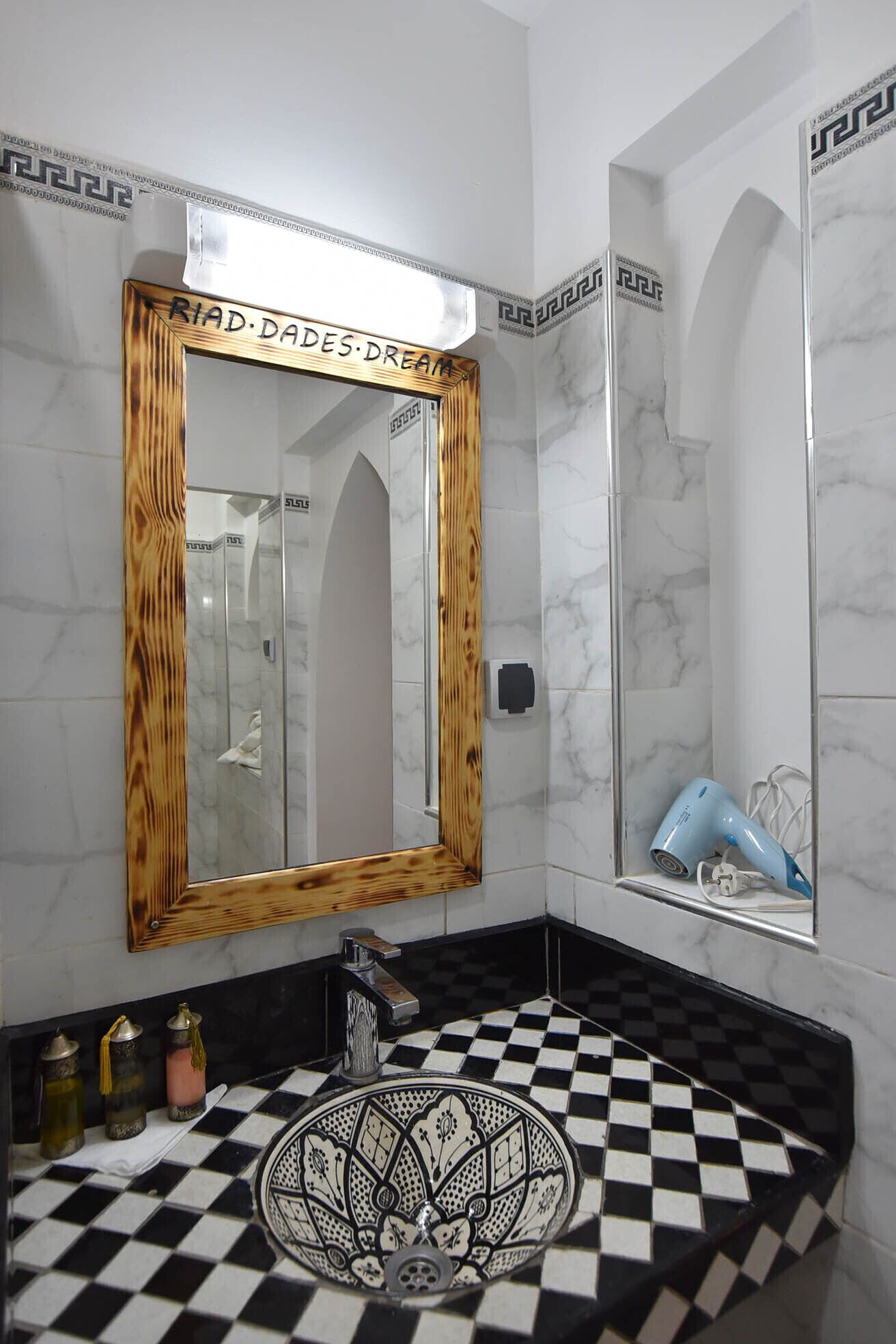 Um banheiro com piso xadrez, pia com cuba decorativa, espelho com moldura de madeira e porta-toalhas com frascos.