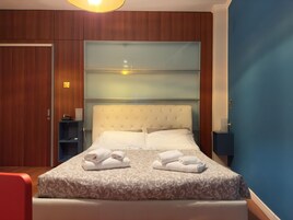 Uma cama bem arrumada com um cobre-leito estampado e duas toalhas dobradas, uma mesa de cabeceira com abajur e um guarda-roupa de madeira.
