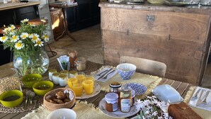 Uma mesa de café da manhã posta com diversos pratos, incluindo frutas, pão e bebidas, em um quarto com um armário de madeira e objetos decorativos.