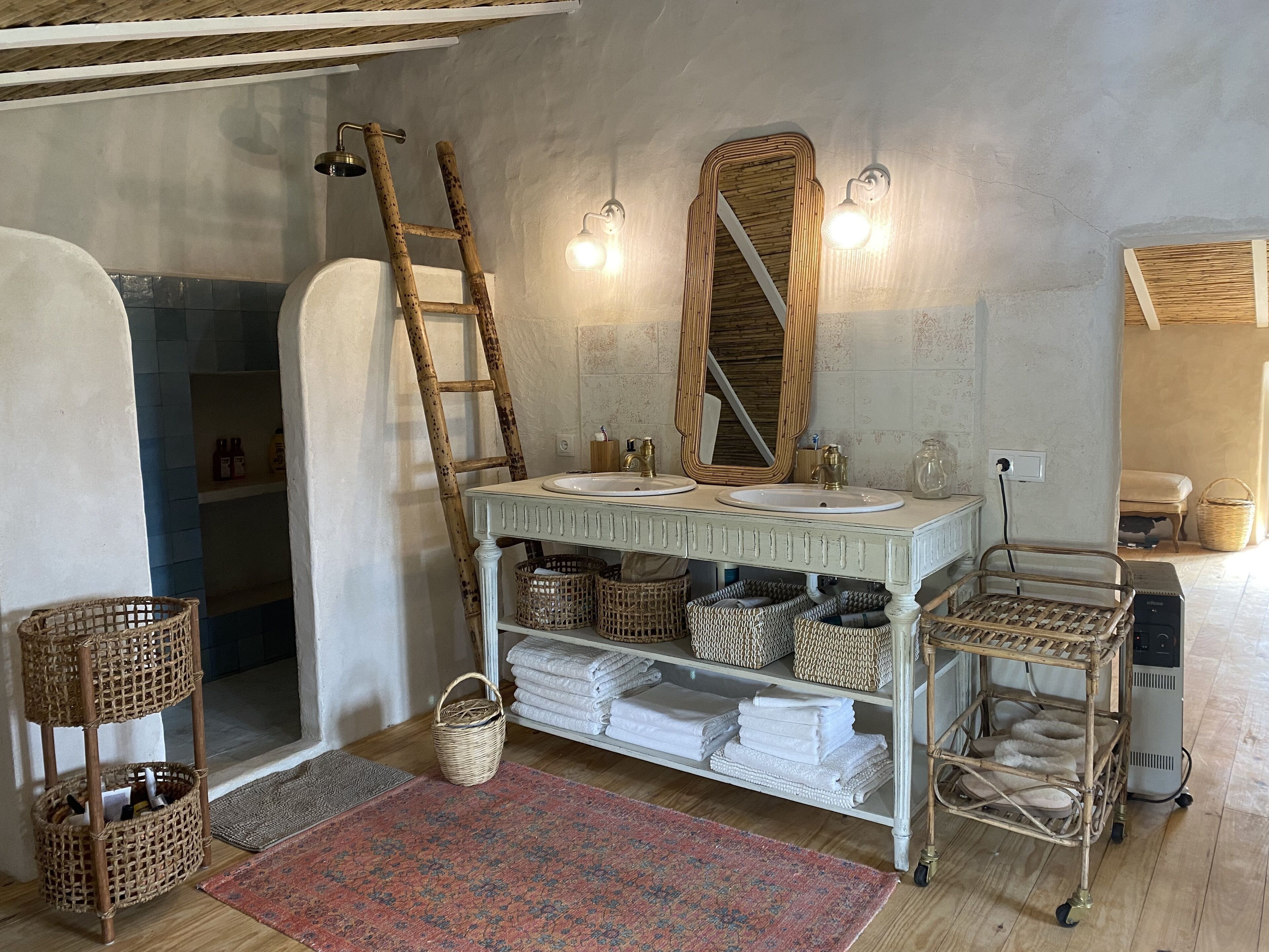 Casa de banho com toucador branco, duas pias, espelho e prateleira com toalhas e cestos.