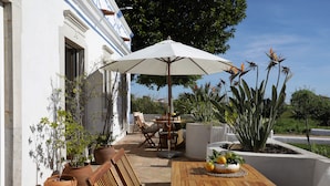 Um pátio externo com mesa e cadeiras de madeira, um guarda-sol branco e plantas em vasos.