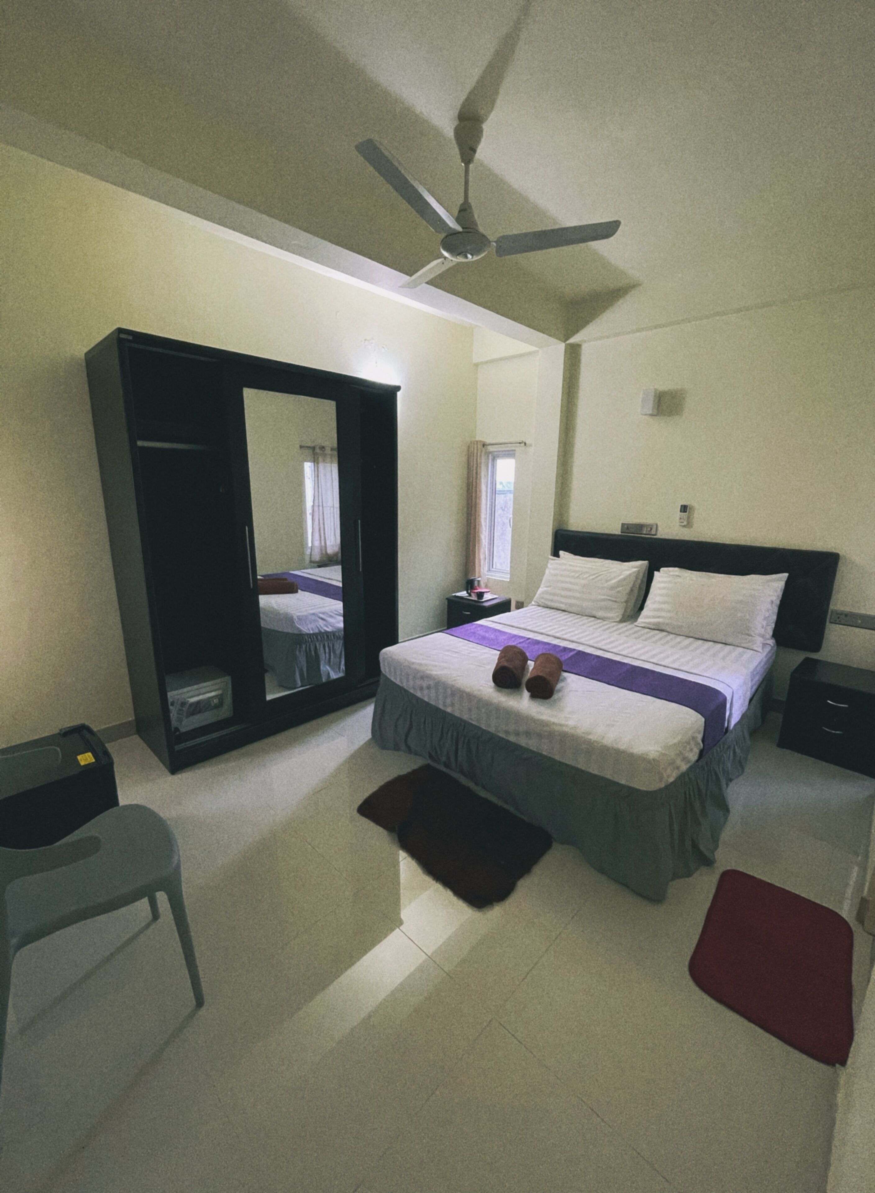 Quarto com cama, guarda-roupa, ventoinha no teto, mesa de cabeceira, cadeira e janela.