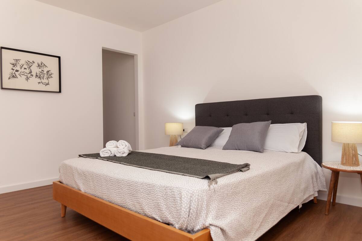 Uma cama bem arrumada com cabeceira cinza, duas luminárias de cabeceira e uma obra de arte emoldurada na parede.