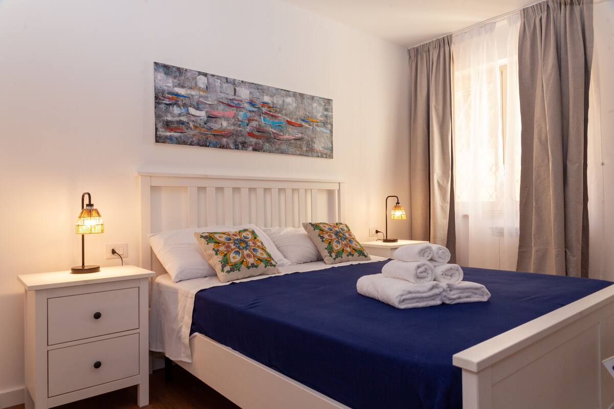 Uma cama bem arrumada com colcha azul, almofadas brancas e travesseiros decorativos, uma luminária de cabeceira e um quadro na parede.
