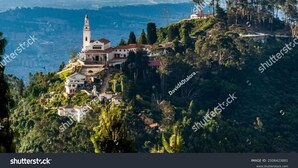 Paisagem montanhosa com uma igreja destacada, apresentando um campanário, rodeada por vegetação exuberante e edifícios residenciais.