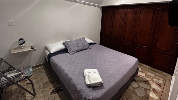 Quarto com cama, guarda-roupa de madeira, mesa de cabeceira pequena com espelho e uma cadeira.