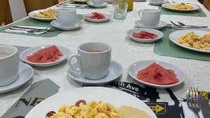 Mesa de café da manhã posta com ovos mexidos, torradas e melancia, além de xícaras e uma televisão ao fundo.