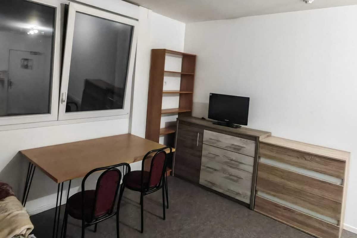 Quarto pequeno e compacto com uma mesa de jantar de madeira e cadeiras, uma TV sobre um móvel de madeira, e uma janela com vidro jateado.
