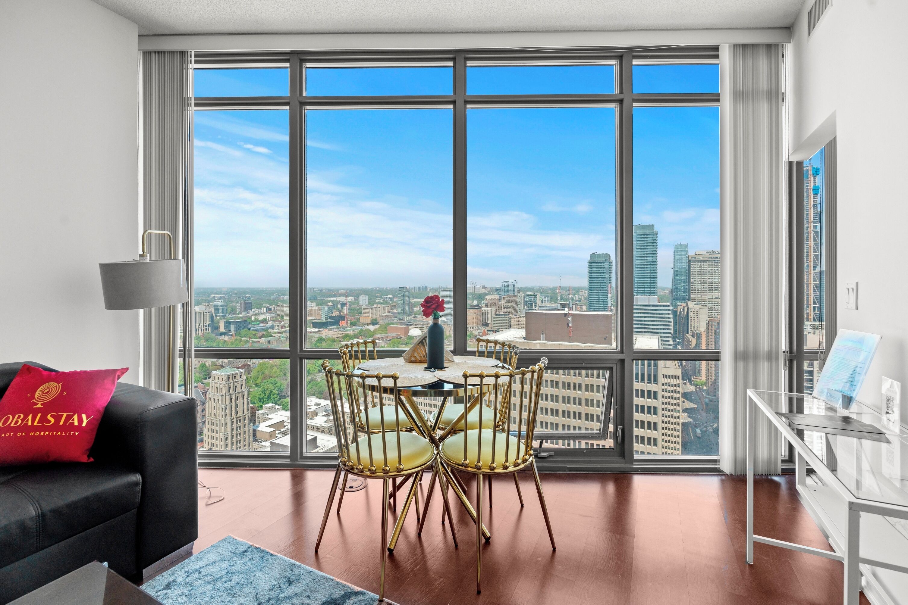 Quarto de hotel moderno com uma janela grande que oferece vista da cidade, um sofá preto com travesseiro vermelho, uma mesa de jantar com cadeiras e uma escrivaninha de vidro.