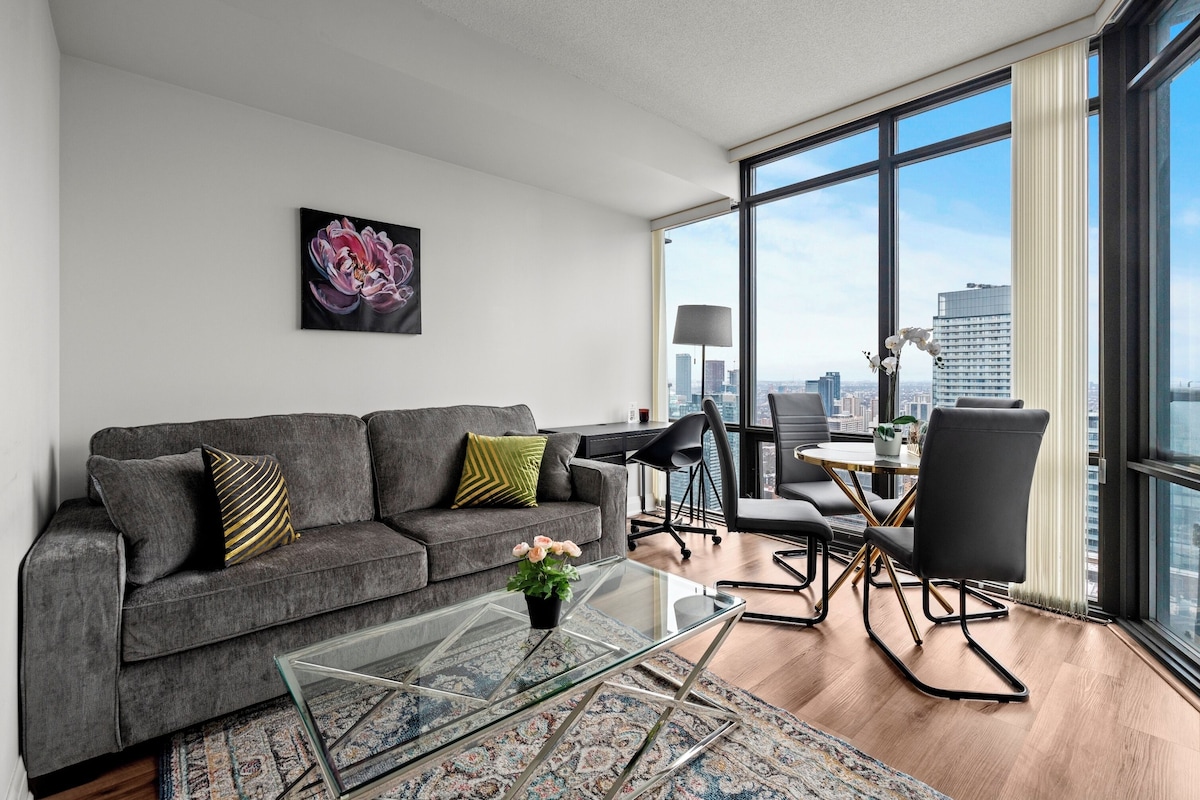 Sala de estar moderna com sofá cinza, mesa de centro de vidro e vista da cidade através de grandes janelas.