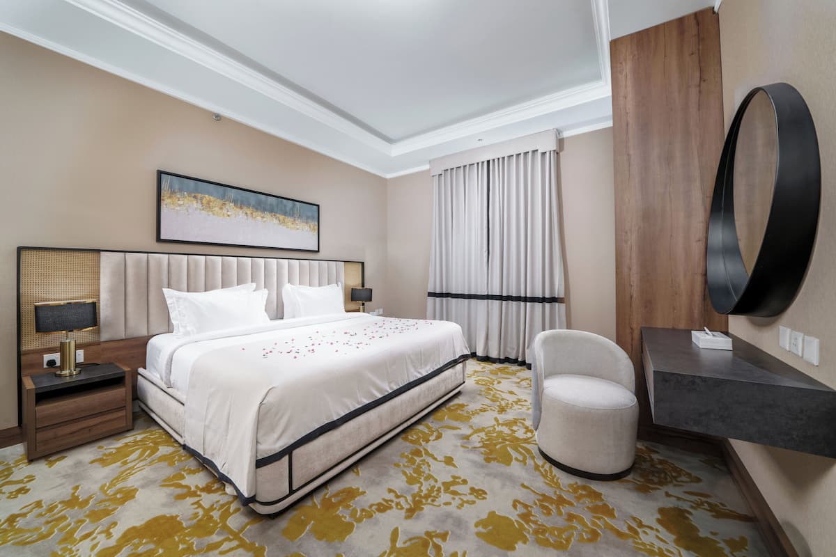 Quarto de hotel com uma cama grande, uma mesa de cabeceira com abajur, uma cadeira, uma escrivaninha com espelho e um quadro na parede.