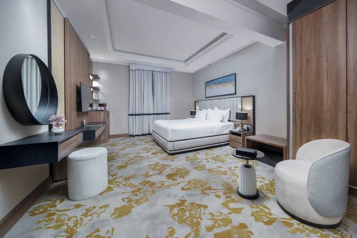Quarto de hotel com uma cama grande, mesa de cabeceira com luminária, cadeira, espelho redondo, TV e parede de painéis de madeira.