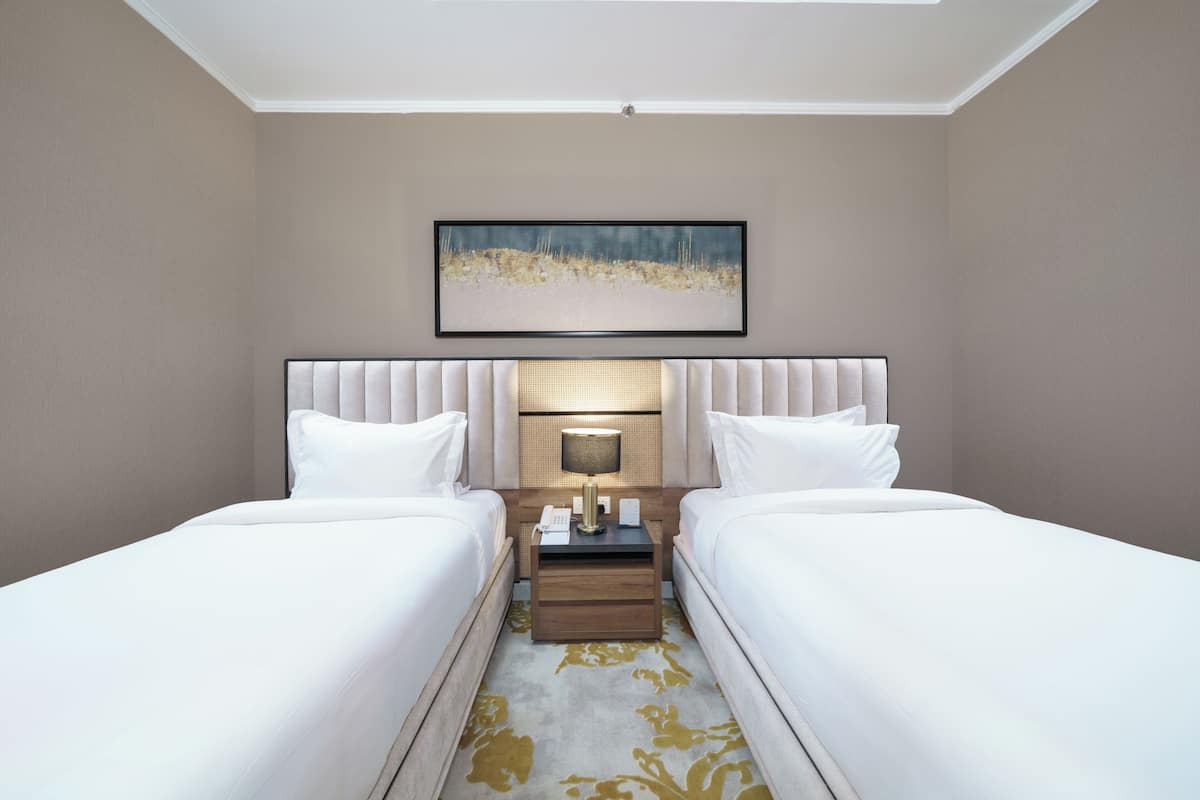 Quarto de hotel com duas camas, uma mesa de cabeceira com abajur e um quadro na parede.