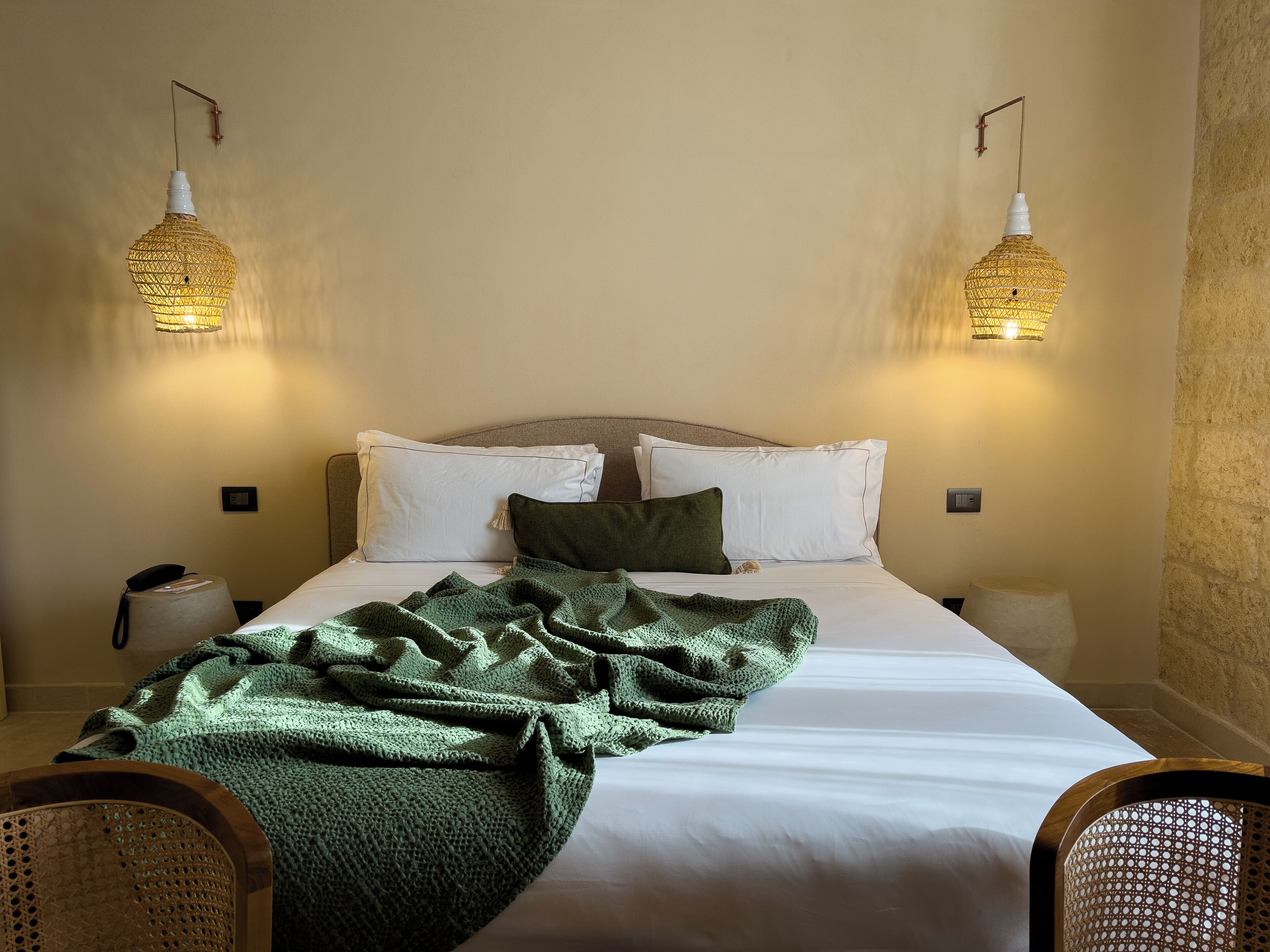 Cama bem arrumada com travesseiros brancos e verdes, manta verde e duas luminárias fixadas na parede.