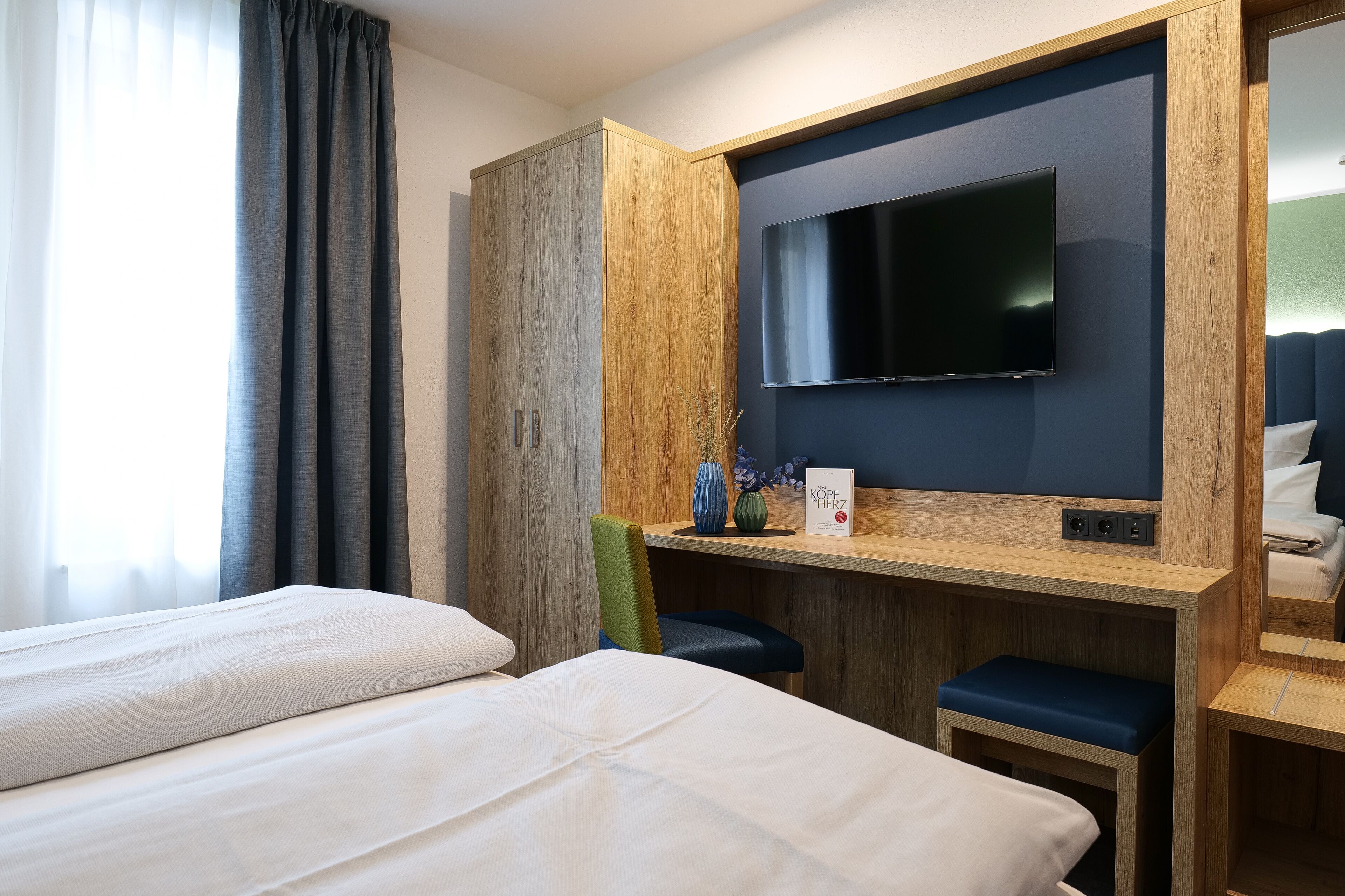 Quarto de hotel com duas camas, um guarda-roupa de madeira, uma televisão na parede e uma pequena mesa com um vaso e uma caixa.