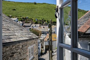 Uma rua estreita com edifícios de pedra, uma colina ao fundo e uma vista através de uma janela com moldura de metal.