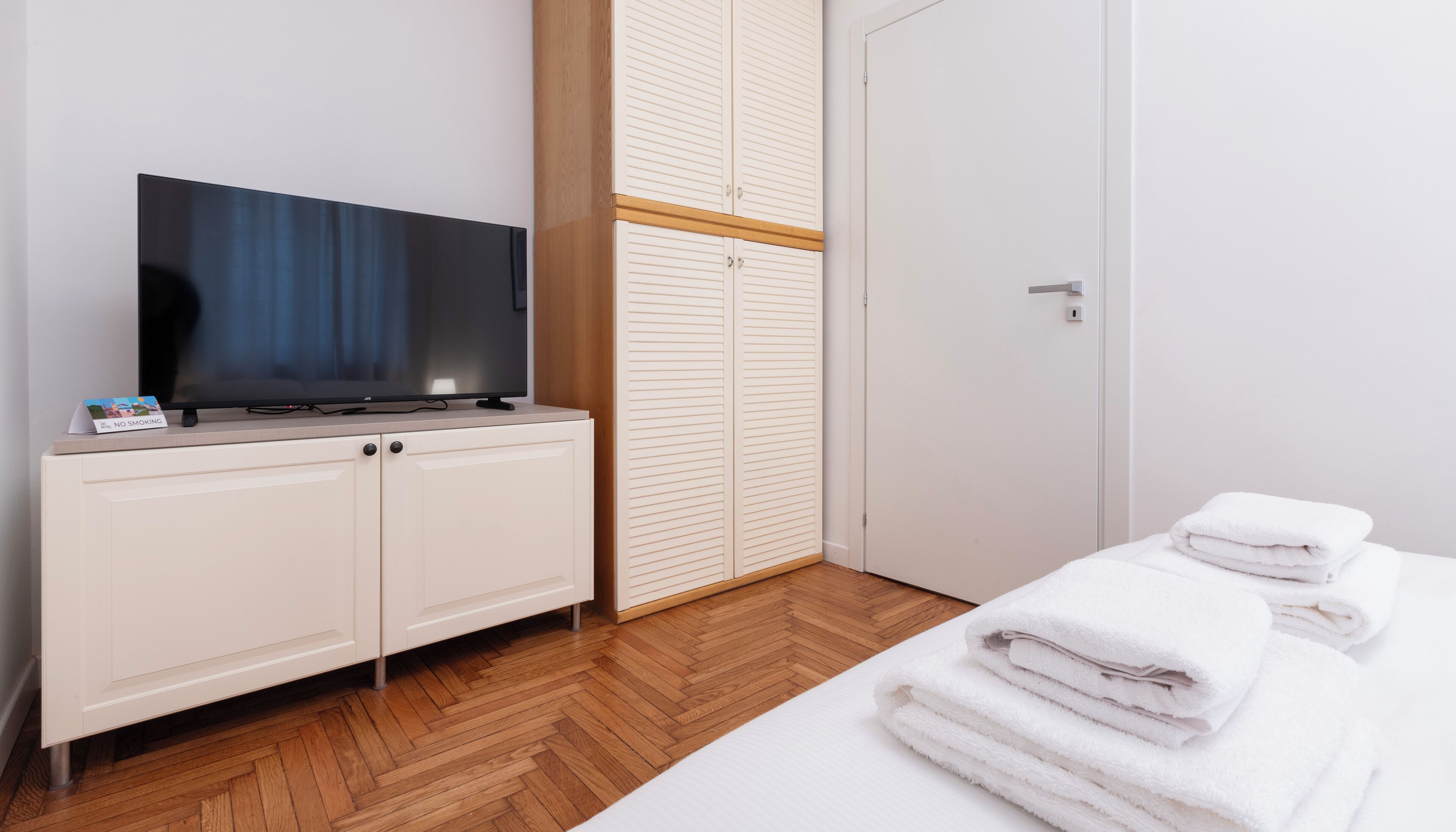 Quarto de hotel com televisão de tela plana em um armário, guarda-roupa com portas deslizantes e cama com toalhas brancas dobradas com cuidado.