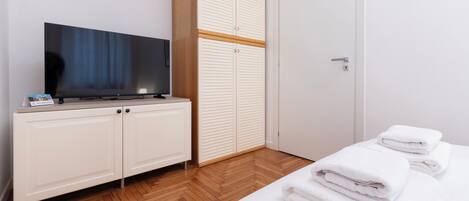 Quarto de hotel com televisão de tela plana em um armário, guarda-roupa com portas deslizantes e cama com toalhas brancas dobradas com cuidado.