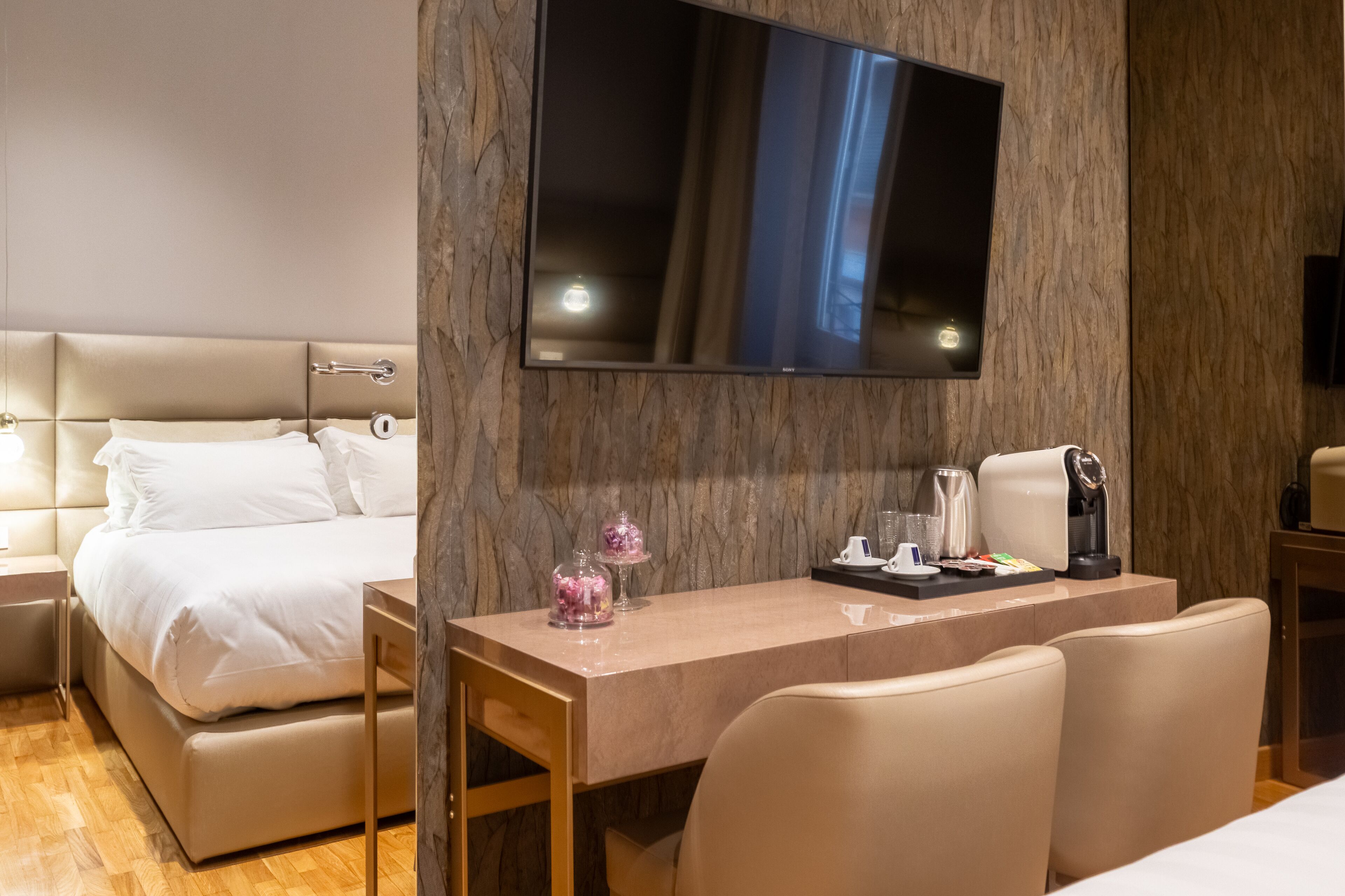 Quarto de hotel com cama, secretária com cafeteira e televisão na parede.