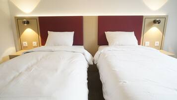 Quarto de hotel com duas camas, cada uma com roupa de cama branca e cabeceira vermelha. Existem luminárias fixadas na parede, uma de cada lado das camas.