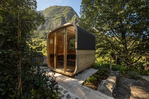 Uma cabana de madeira moderna e arredondada com grandes janelas de vidro, situada num jardim exuberante com montanhas ao fundo.