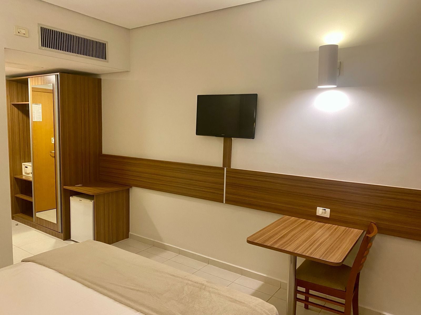 Quarto de hotel com cama, uma pequena mesa, televisão fixada na parede e armário com espelho.