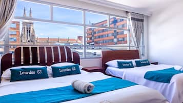 Quarto de hotel com duas camas, cada uma com colcha e almofadas azuis, identificadas como 'Lourdes 62'. O quarto possui janelas amplas com vista para um horizonte urbano, incluindo uma igreja e edifícios modernos.