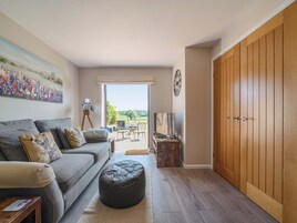 Sala de estar com sofá cinza, um banquinho e um quadro de flores. Há um guarda-roupa de madeira e uma porta que dá para o exterior.