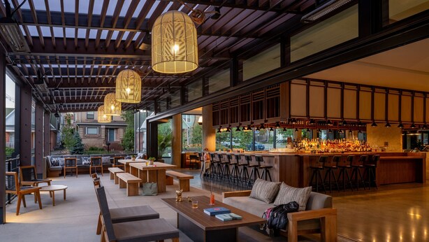 Uma área externa ampla com mesas e cadeiras de madeira, um bar com banquetas e luminárias de vime suspensas.