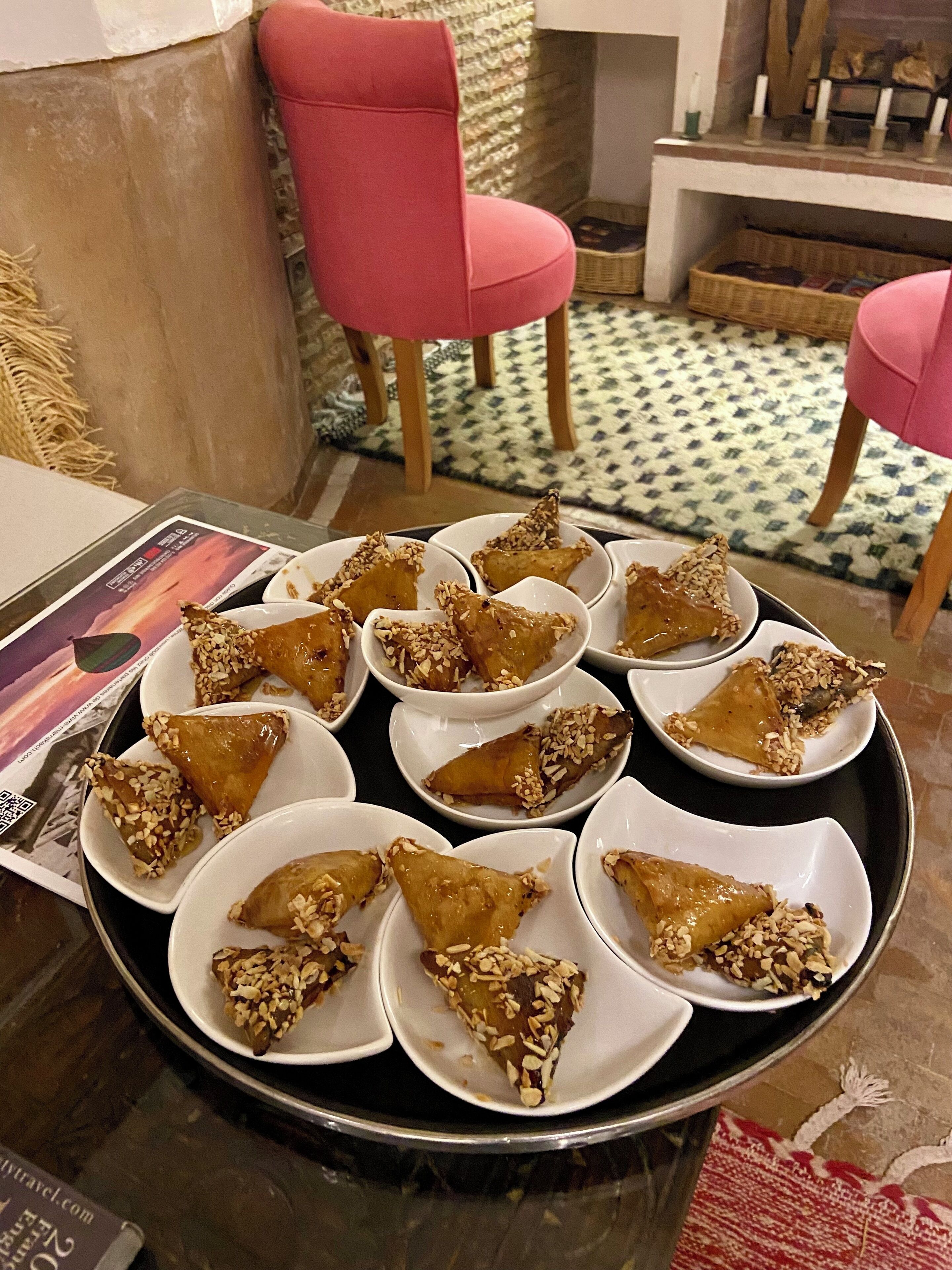 Uma bandeja com pastéis triangulares decorados com frutos secos, colocada sobre uma mesa num espaço com chão estampado e lareira de tijolos.