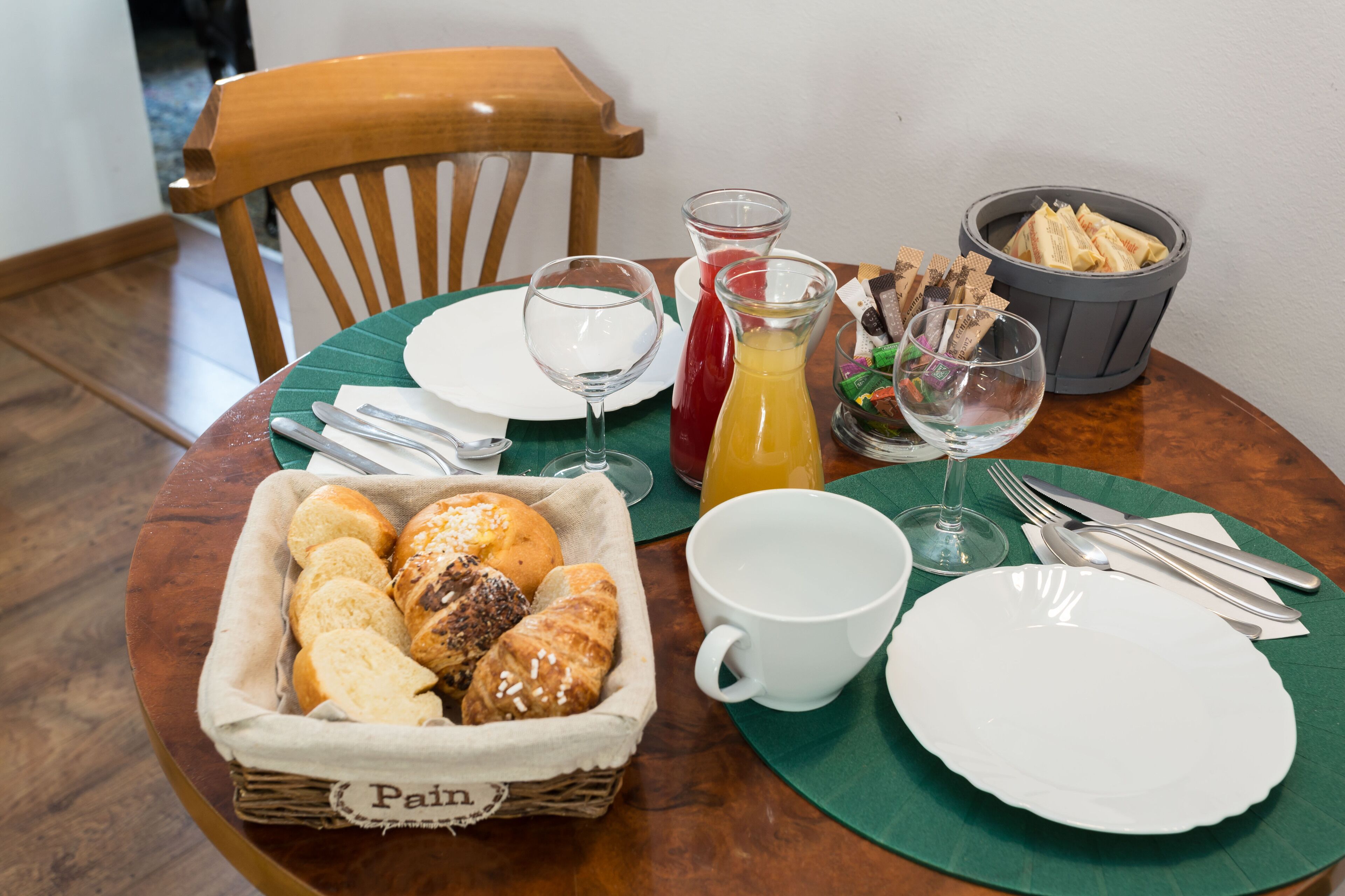 Disposição de café da manhã com uma cesta de pães e doces, um jarro de suco de laranja e um copo de suco vermelho sobre uma mesa de madeira.
