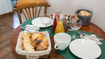 Disposição de café da manhã com uma cesta de pães e doces, um jarro de suco de laranja e um copo de suco vermelho sobre uma mesa de madeira.