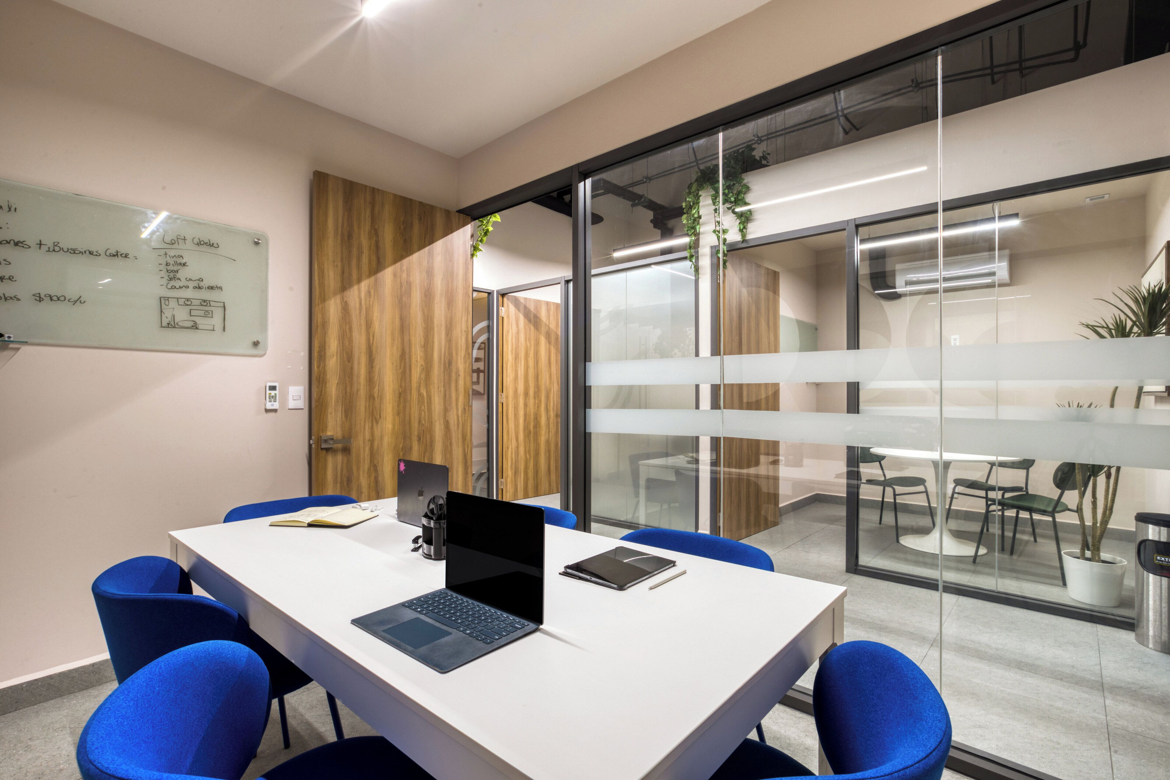 Uma sala de reunião moderna com uma mesa branca, cadeiras azuis, um laptop e um quadro branco com texto.