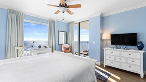 Quarto com uma cama grande, ventilador de teto, televisão e vista para o mar.