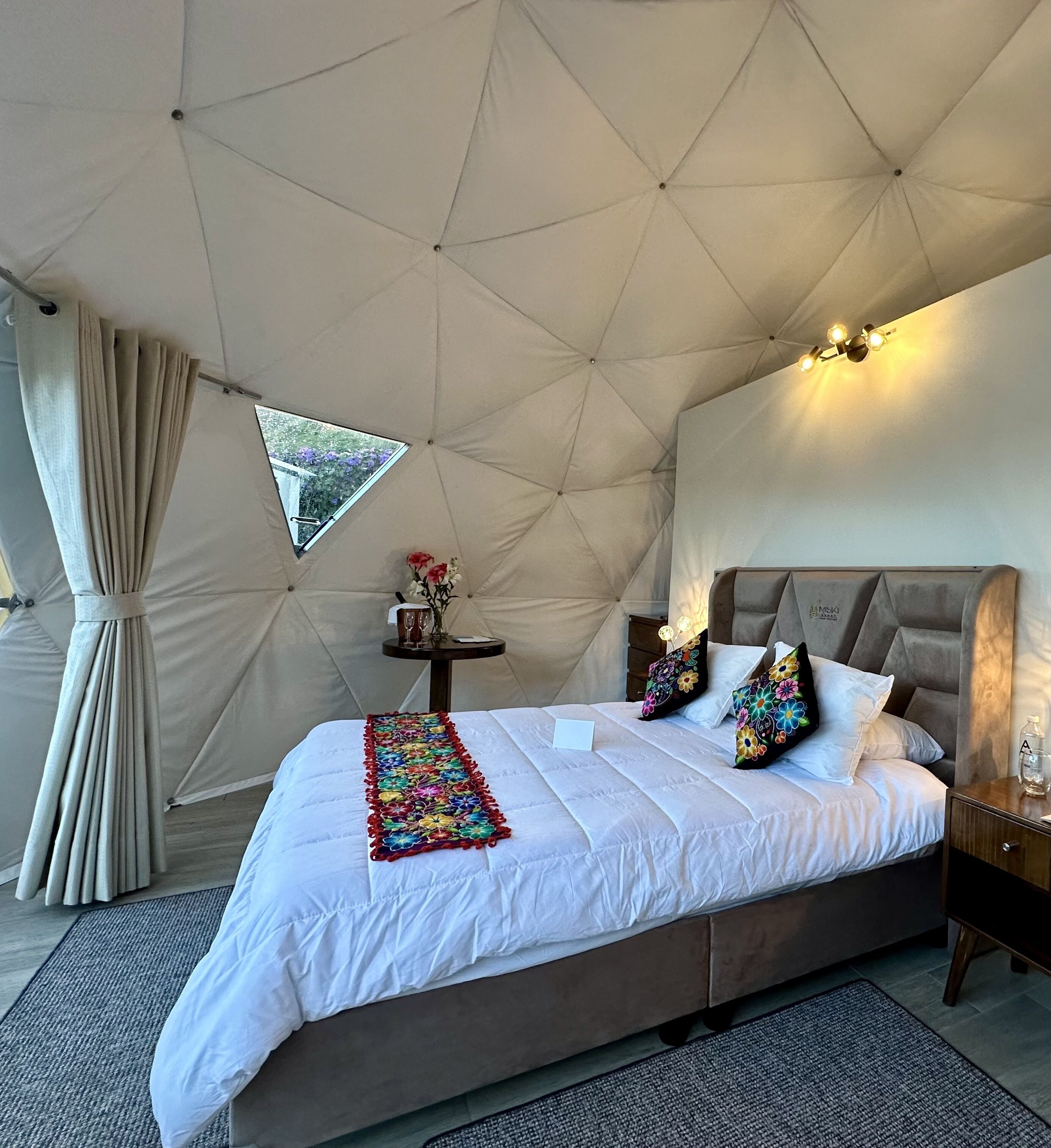 Quarto com uma cama grande, uma mesa pequena com um vaso e vista para o exterior por uma janela triangular.