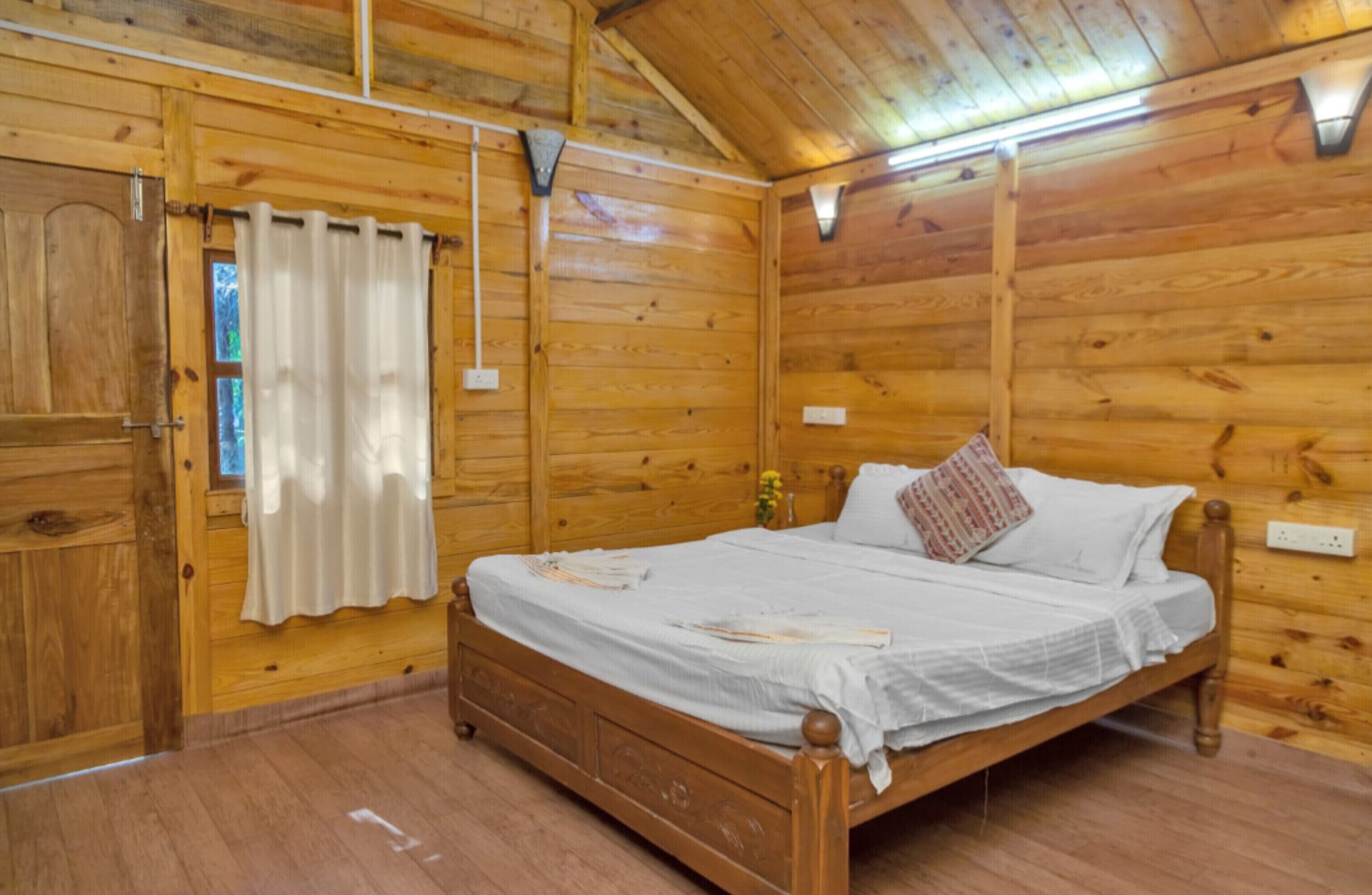 Quarto de cabana de madeira com uma cama de solteiro, uma porta de madeira e uma janela com cortinas.