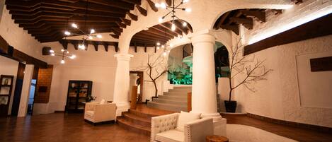 Um hall espaçoso com teto alto, vigas de madeira e uma escadaria que leva a um andar superior. Há colunas brancas e um sofá branco no centro.